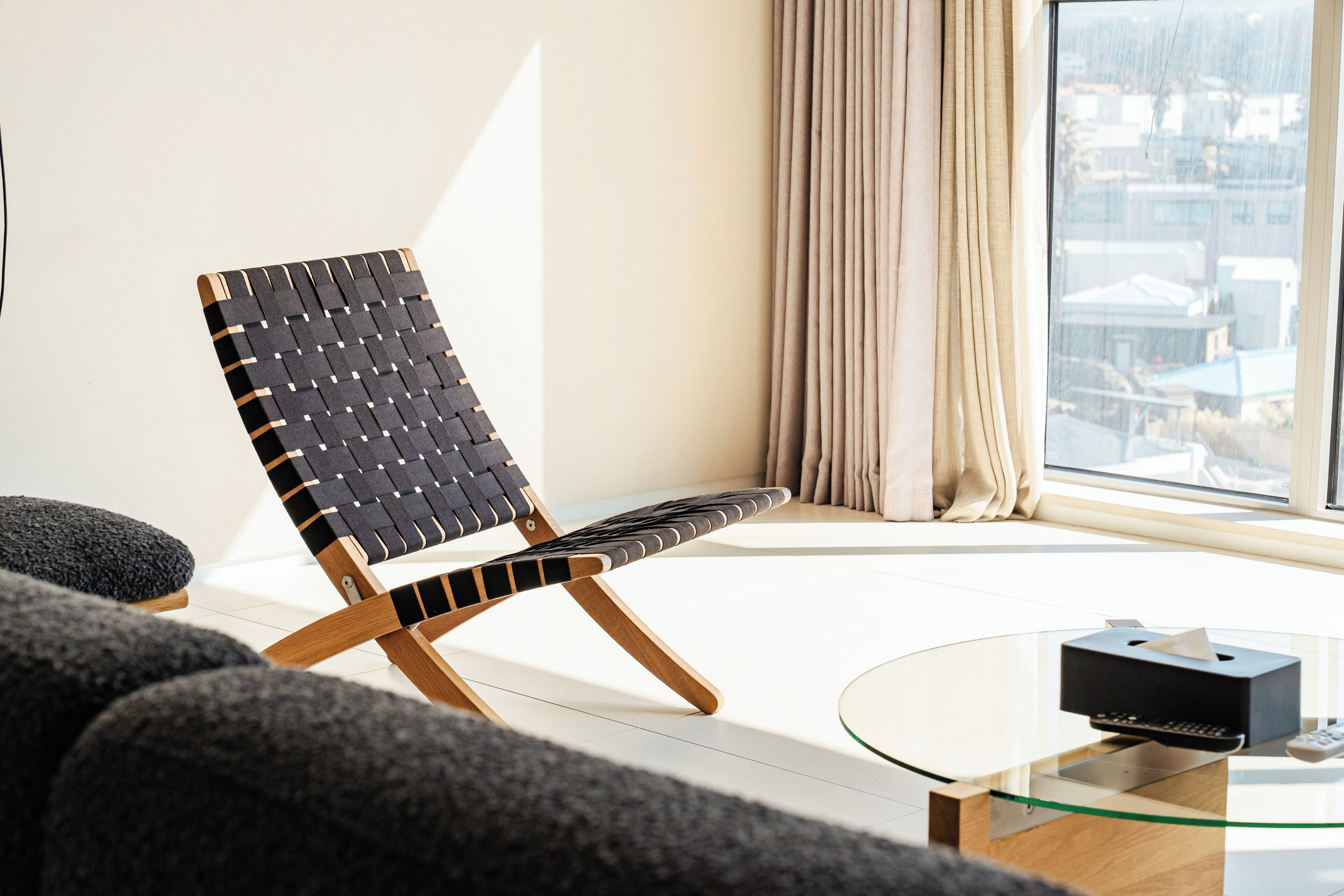 Sala de estar moderna com uma cadeira trançada preta, uma mesa de centro de vidro e vista para um horizonte urbano através de cortinas bege.
