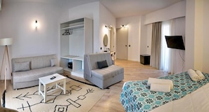 Quarto de hotel moderno com sofá, uma pequena mesa, cadeira, cama com colcha estampada, televisão e guarda-roupa.