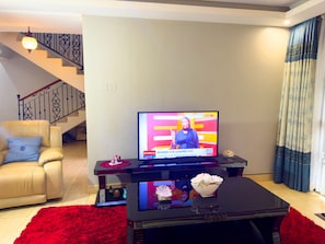 Uma sala de estar com televisão, sofá, mesa de centro com objetos decorativos e uma escada ao fundo.