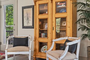 Um quarto com duas cadeiras antigas brancas, com assentos e encostos acolchoados, um armário de madeira com portas de vidro e um quadro na parede.
