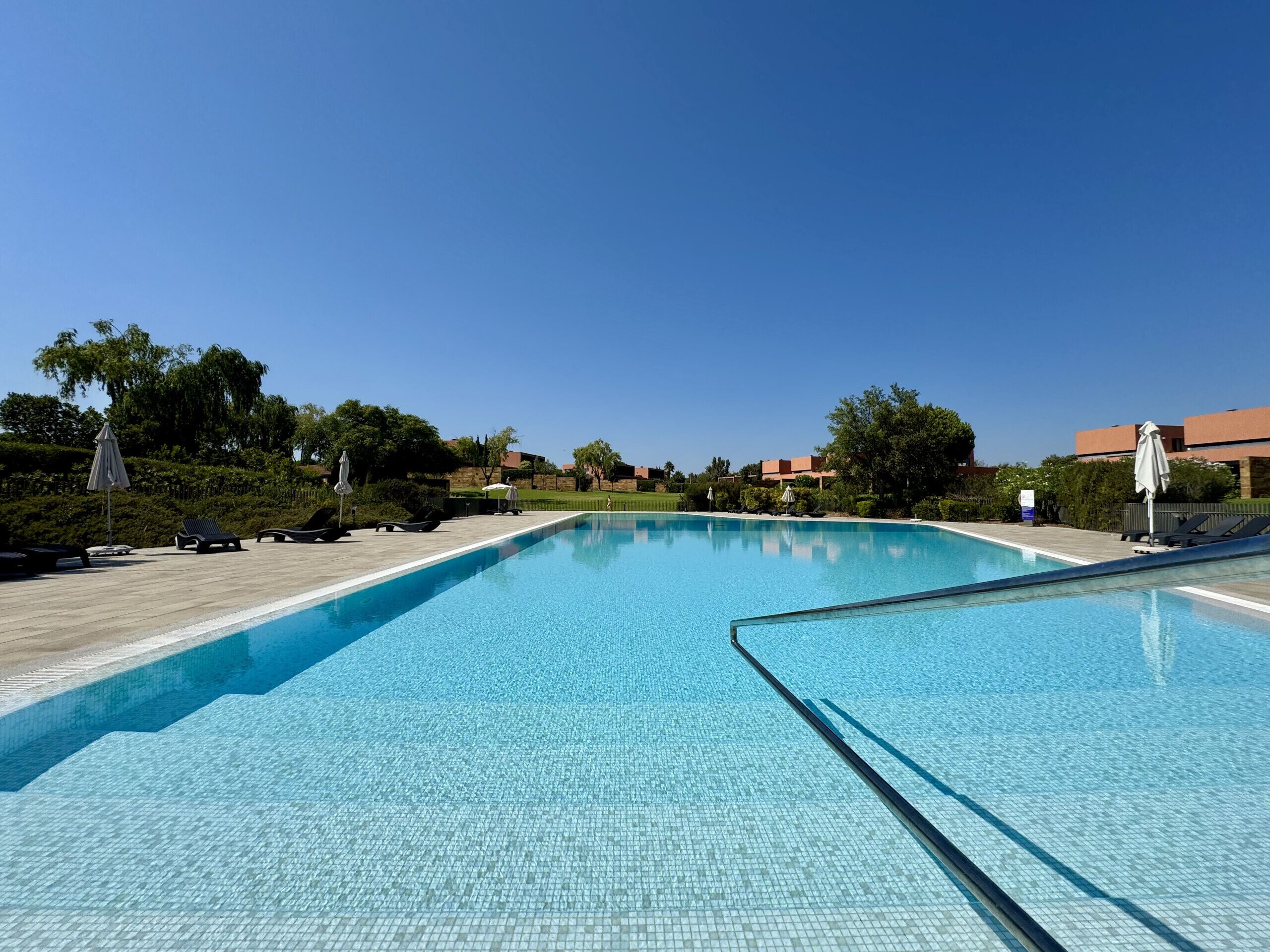 Piscina azul límpida com espreguiçadeiras e guarda-sóis, cercada por vegetação e edifícios.