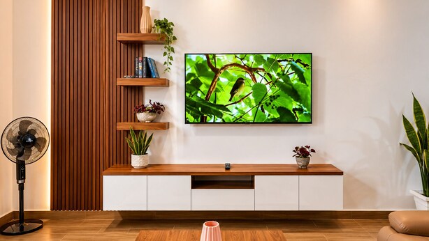 Uma sala de estar moderna com televisão de tela plana na parede, prateleiras de madeira, um vaso de planta, um ventilador de chão e uma mesa de centro.