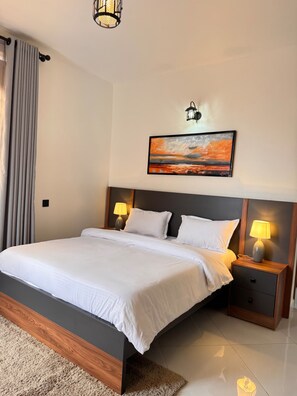 Uma cama bem arrumada com lençóis brancos, cabeceira de madeira, duas mesas de cabeceira com abajures, um quadro sobre a cama e uma cortina cinza na janela.