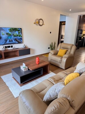 Uma sala de estar moderna com televisão de tela plana, uma mesa de centro de madeira, um sofá bege e uma poltrona de couro.