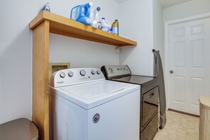 Uma lavanderia com máquina de lavar e secadora Whirlpool, uma prateleira de madeira com produtos de limpeza e uma porta com porta-toalhas.