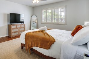 Quarto com uma cama, televisão sobre um armário de madeira, janela com persianas e ventilador de teto.