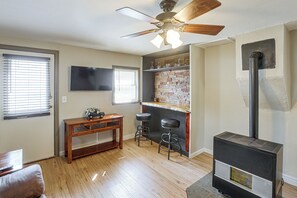 Uma sala de estar com piso de madeira, ventilador de teto, lareira, TV fixada na parede e uma janela com persianas.