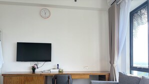 Quarto de hotel com cama, sofá, escrivaninha com televisão de tela plana, uma cadeira, relógio na parede e janela com cortinas.