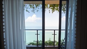 Quarto com cama, vista para o mar através de uma porta de vidro e varanda com plantas.