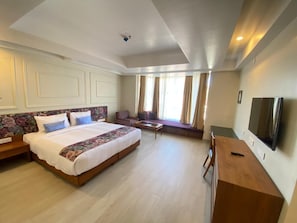 Quarto de hotel com cama grande, televisão, área de estar e janela com cortinas.