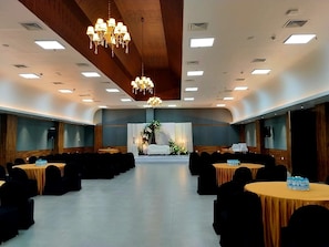 Uma sala de conferências com mesas redondas, cadeiras pretas, um palco com decorações florais e um candelabro.