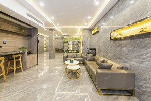 Um lobby moderno de hotel com piso de mármore, área de bar, assentos com sofá e poltronas, e uma parede decorativa com plantas.