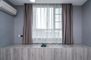 Quarto com uma grande janela, cortinas cinzas, assoalho de madeira e um ar condicionado.