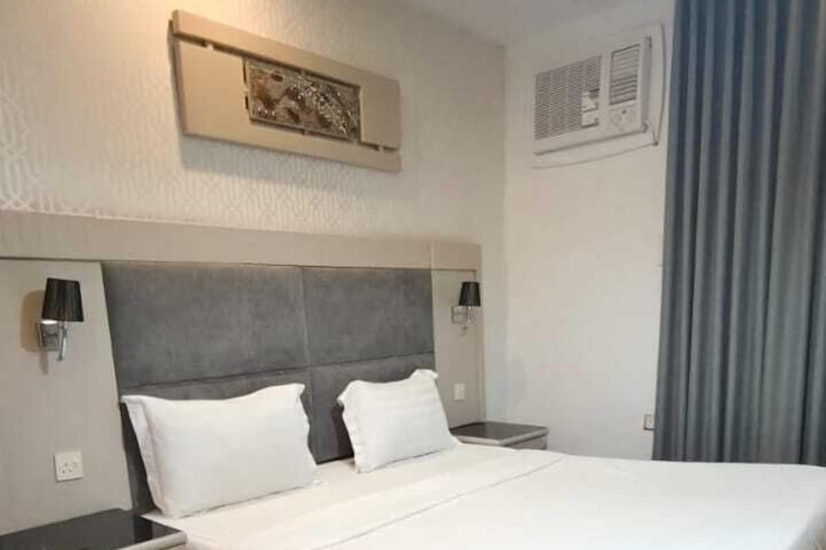 Quarto de hotel com uma cama grande, mesas de cabeceira, cabeceira, luminárias fixadas na parede, iluminação de teto e uma janela com cortinas.