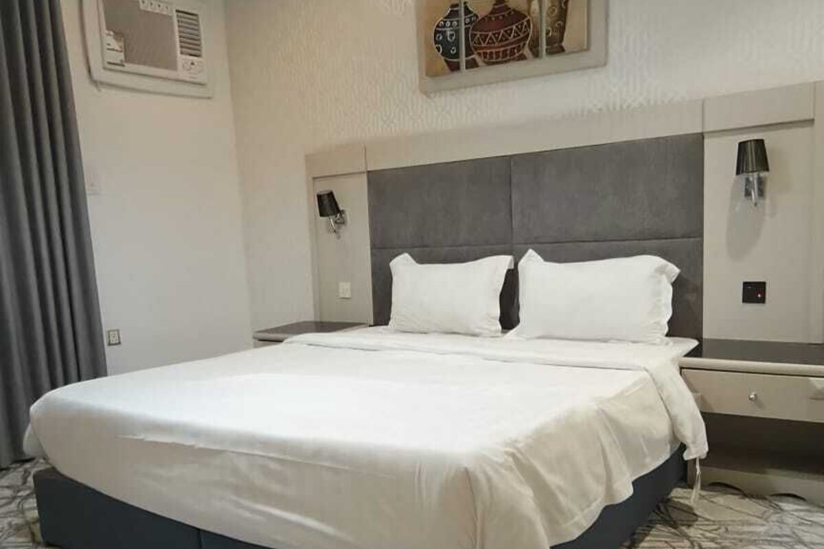 Quarto de hotel com cama grande, mesa de cabeceira, abajur e uma obra de arte na parede.