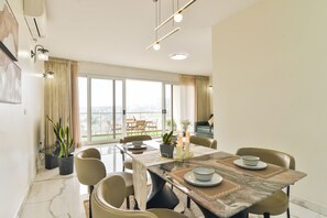 Uma área de refeições com uma mesa posta para quatro pessoas, cadeiras, plantas em vasos e vista da cidade através de grandes janelas.