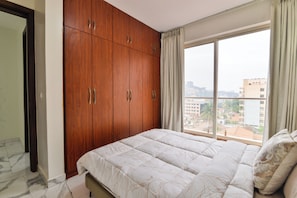 Quarto com um guarda-roupa espaçoso, cama com colcha estampada e vista para a cidade pela janela.