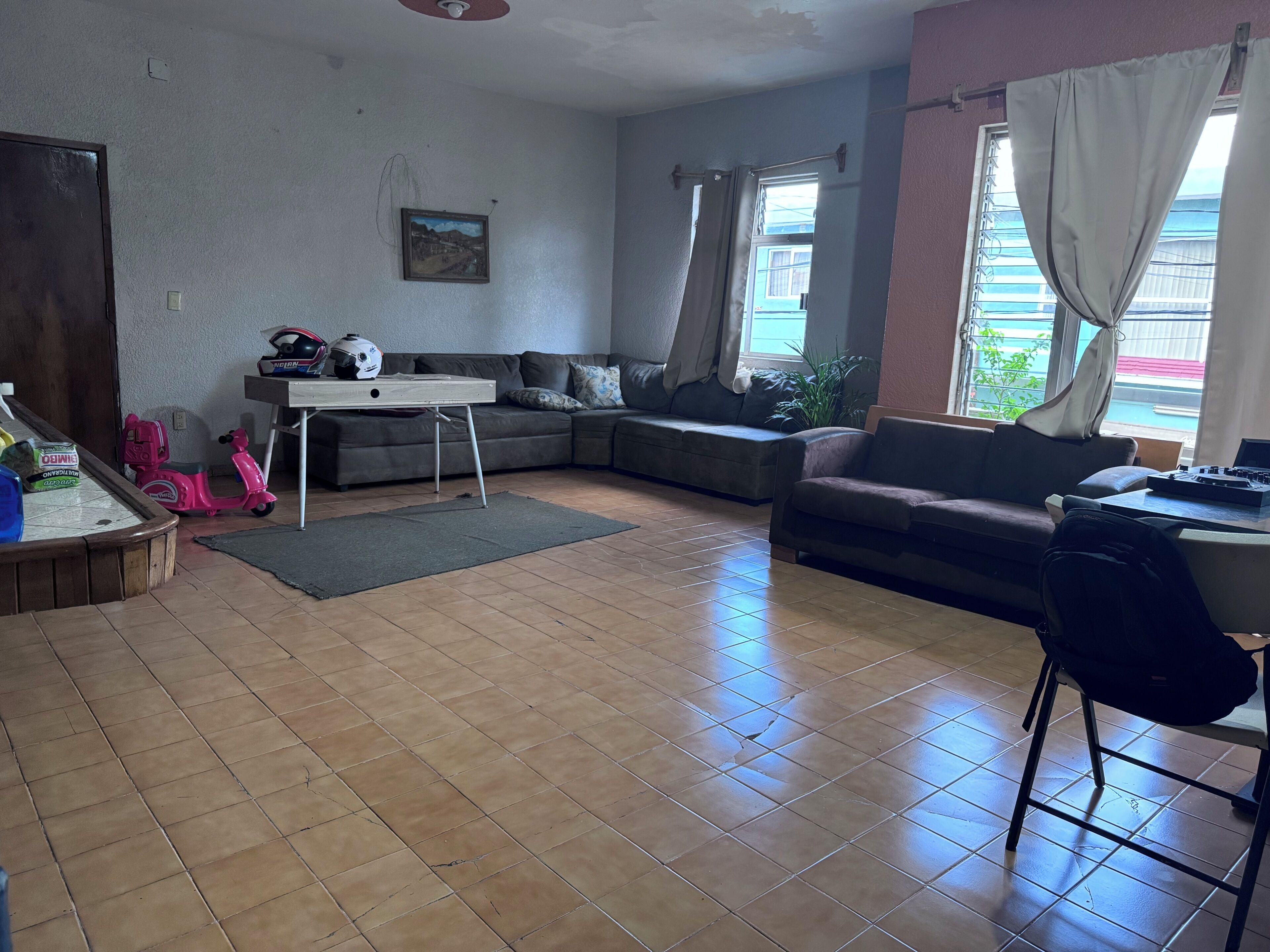 Uma sala de estar com sofá, mesa de centro e televisão. Há uma janela com cortinas e uma planta.