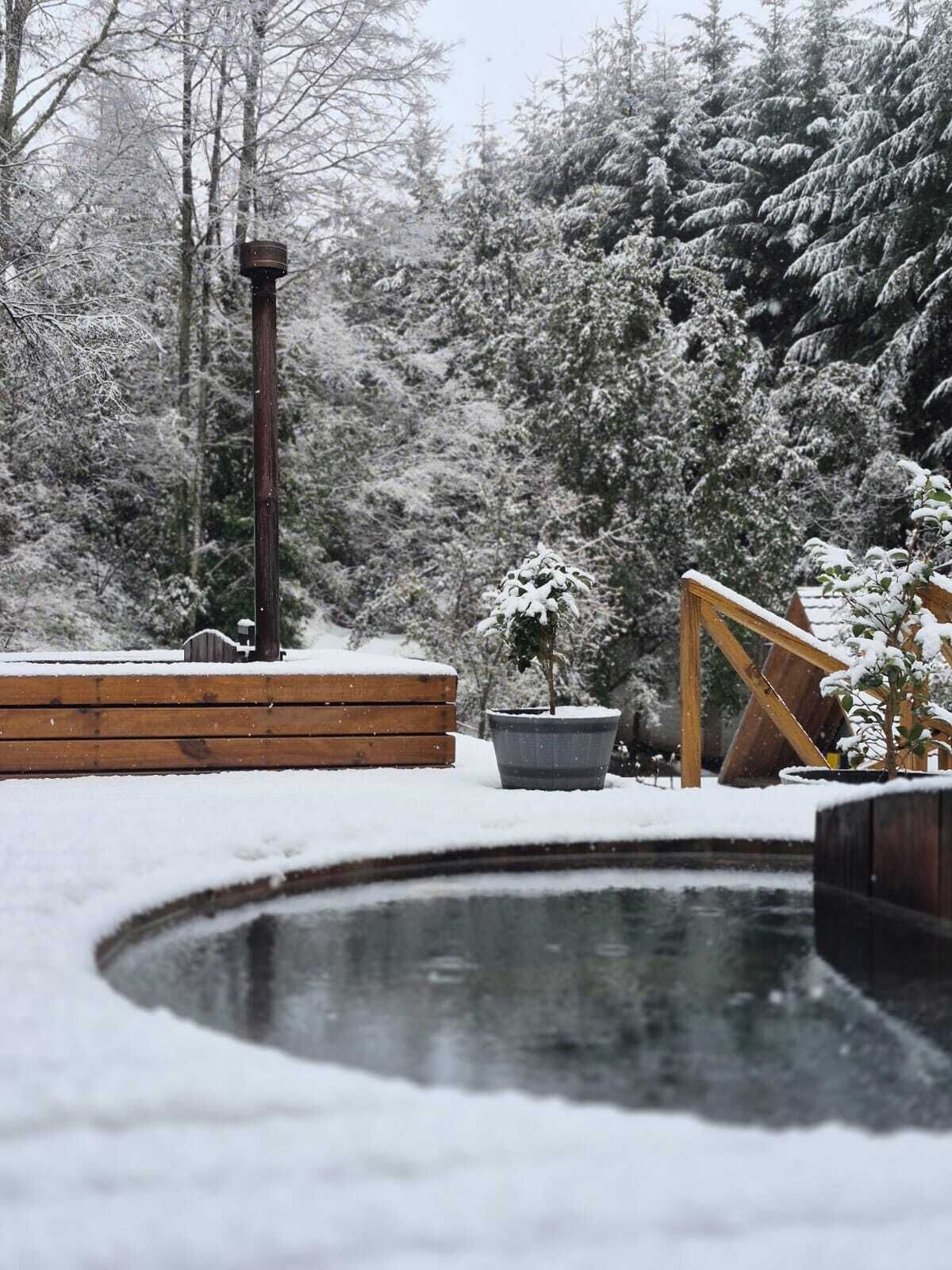 Área externa coberta de neve, com um deck de madeira, estrutura metálica e um pequeno lago.