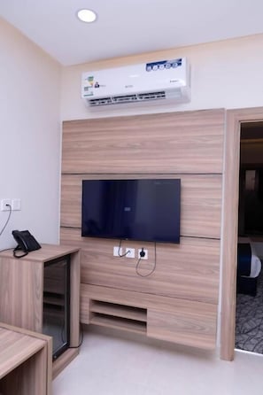 Quarto com televisão de tela plana montada, paredes de painel de madeira, unidade de ar condicionado embutida e uma pequena escrivaninha com telefone.