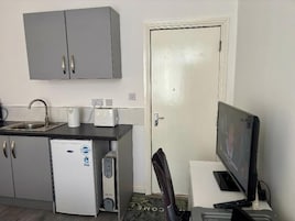 Uma cozinha compacta com geladeira, pia e micro-ondas. Há uma porta com puxador e uma televisão sobre um suporte.