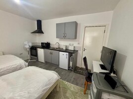 Quarto de hotel compacto com duas camas de solteiro, uma kitchenette, uma televisão e uma pequena mesa de trabalho.