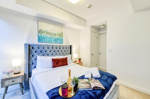Cama bem arrumada com cabeceira estofada, mesa de cabeceira com abajur, uma garrafa de champanhe e bandeja com um copo e uma vela.