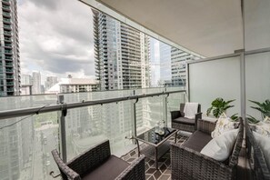 Uma varanda com móveis de vime, guarda-corpo de vidro e vista para a cidade.