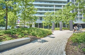 Uma passarela pavimentada com vegetação, bancos e um edifício moderno com vários andares ao fundo.