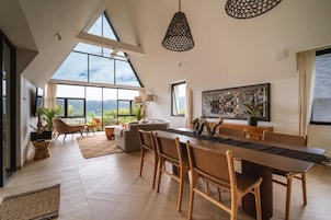 Um quarto espaçoso com teto alto, uma mesa de jantar com cadeiras, um sofá e uma grande janela com vista para o exterior.