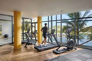 Academia com esteiras, elípticos e uma grande janela com vista para uma piscina e palmeiras.