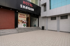 Entrada de hotel com uma persiana fechada, uma placa anunciando 'HOTEL SHAGUN' e um caminho de paralelepípedos que leva à entrada.