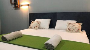 Uma cama bem arrumada com lençóis brancos e manta verde, dois travesseiros decorativos e uma toalha enrolada ao lado.