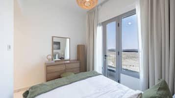 Quarto com cama, penteadeira, espelho e vista da praia por uma grande janela.