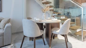 Uma sala de jantar moderna com uma mesa redonda, duas cadeiras cinzas estofadas, um vaso com plantas secas e uma escada com degraus de madeira que dá para uma cozinha.