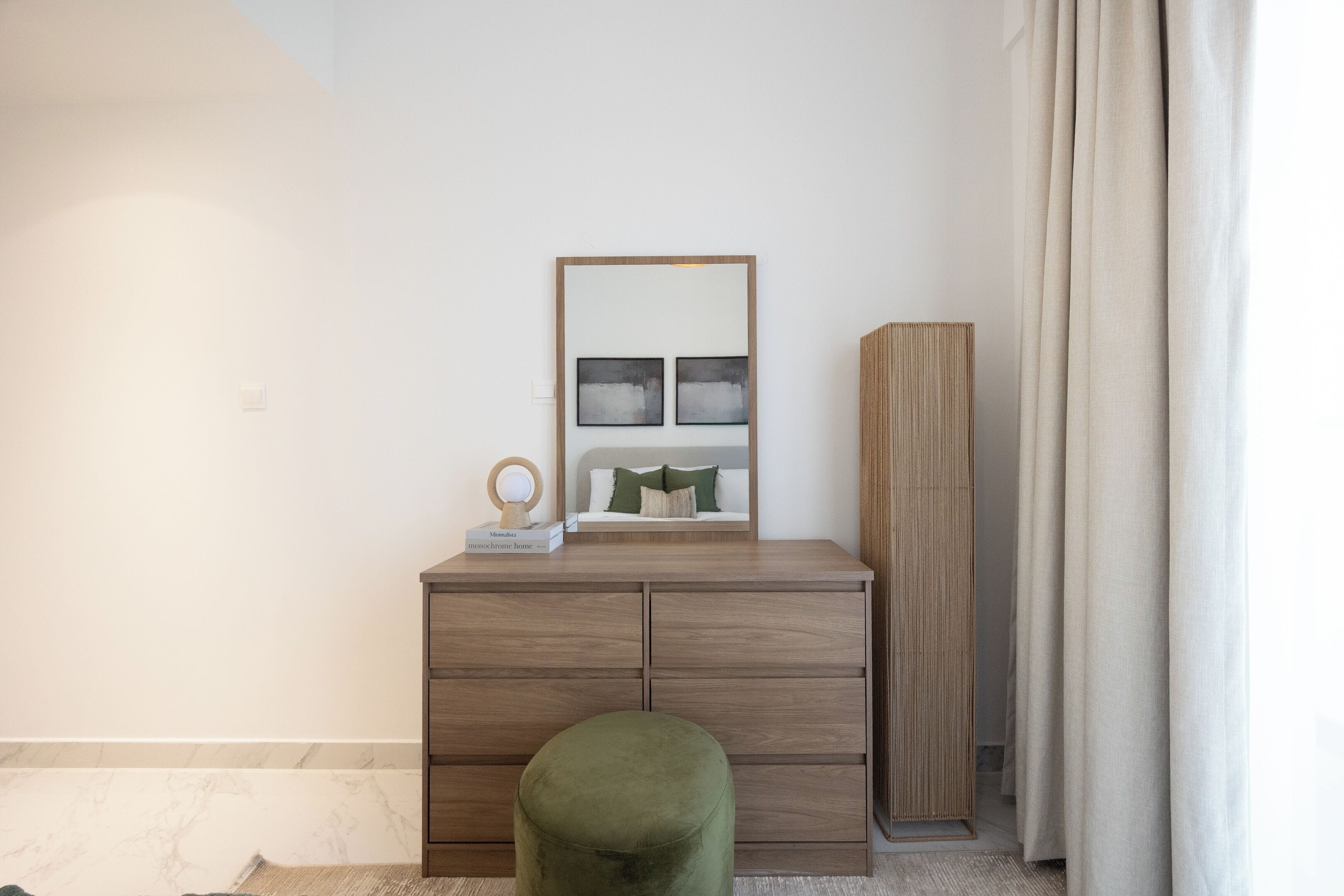 Quarto minimalista com cômoda de madeira, um espelho e um banquinho verde.