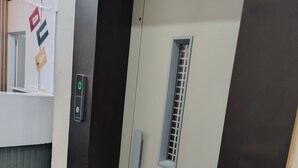 Um elevador moderno com painel digital, uma pequena janela e um balcão de recepção ao fundo.