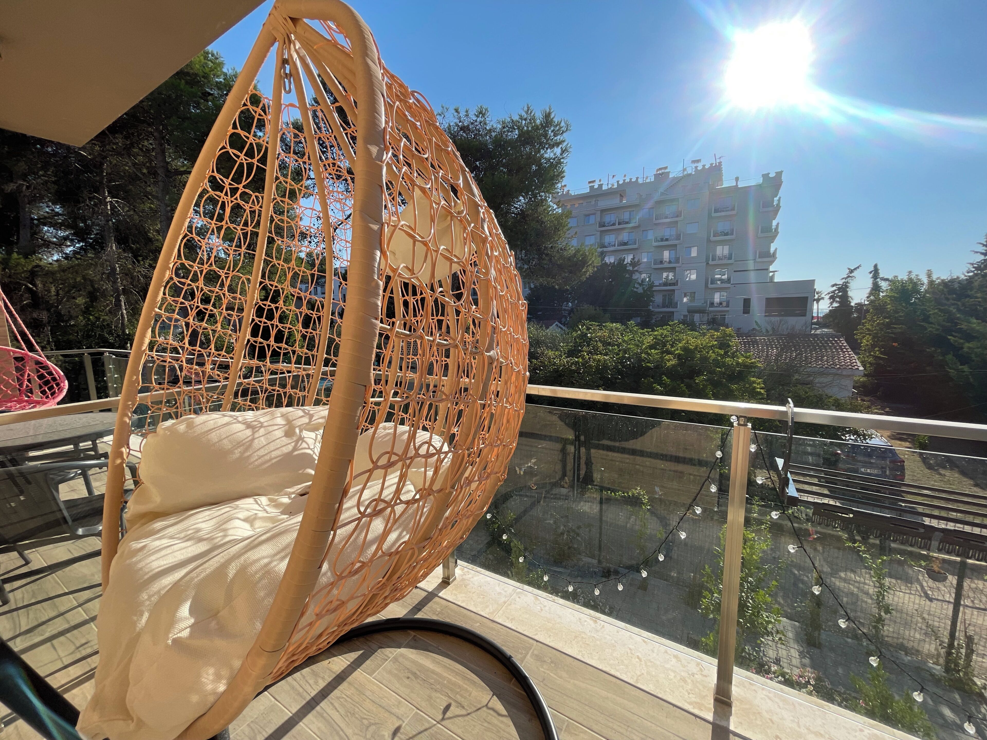 Uma varanda com uma cadeira suspensa, guarda-corpo de vidro e vista para um edifício ao fundo.