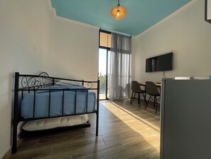 Quarto com cama, escrivaninha, cadeira, televisão e janela com cortinas.