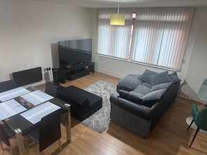 Uma sala de estar moderna com televisão de tela plana, um sofá, uma mesa de jantar com cadeiras e uma janela com persianas.