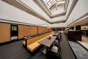 Um espaço amplo e interno, com pé direito alto, área de estar central com bancos e mesas, e um balcão de recepção.