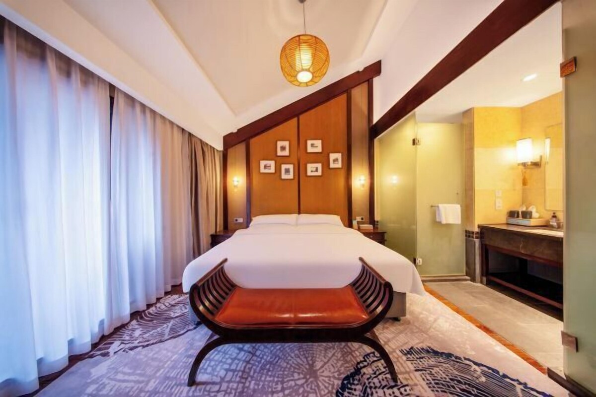Quarto de hotel com cama, um banco de madeira, televisão presa na parede e banheiro com espelho e pia.