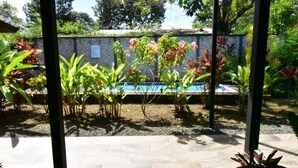 Área de piscina com piso de azulejos, cercada por plantas variadas e uma parede de pedra.