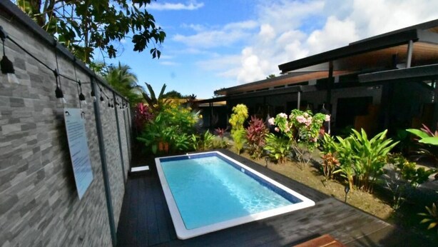 Piscina com deck de madeira, cercada por vegetação exuberante e uma construção moderna.