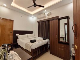 Quarto de hotel com cama, guarda-roupa, escrivaninha com chaleira e ventoinha no teto.