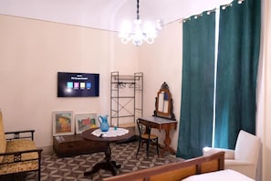 Um quarto com uma cama, uma cadeira, uma mesa com um vaso, uma televisão fixada na parede e uma janela com cortinas.