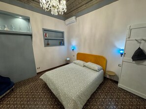 Um quarto com carpete estampado, uma cama com colcha estampada, um lustre, um guarda-roupa e uma pequena mesa com abajur.