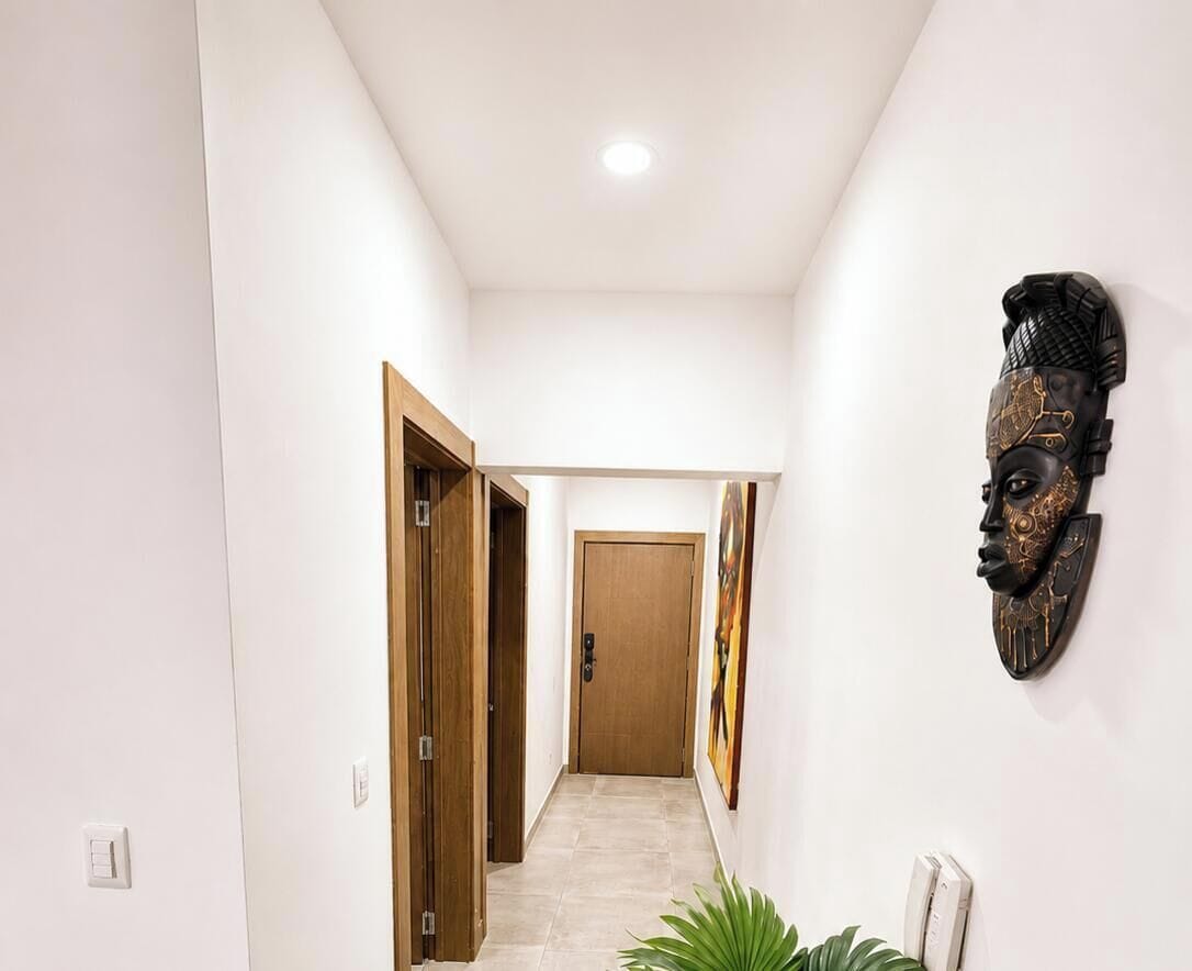 Um corredor com uma porta de madeira no fim, uma máscara decorativa na parede e um vaso com planta.