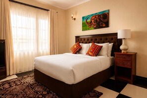 Quarto de hotel com uma cama grande, cabeceira, um quadro, luminária e tapete estampado.