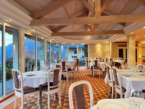 Restaurante com várias mesas e cadeiras, grandes janelas com vista para o exterior e estrutura de teto em madeira.